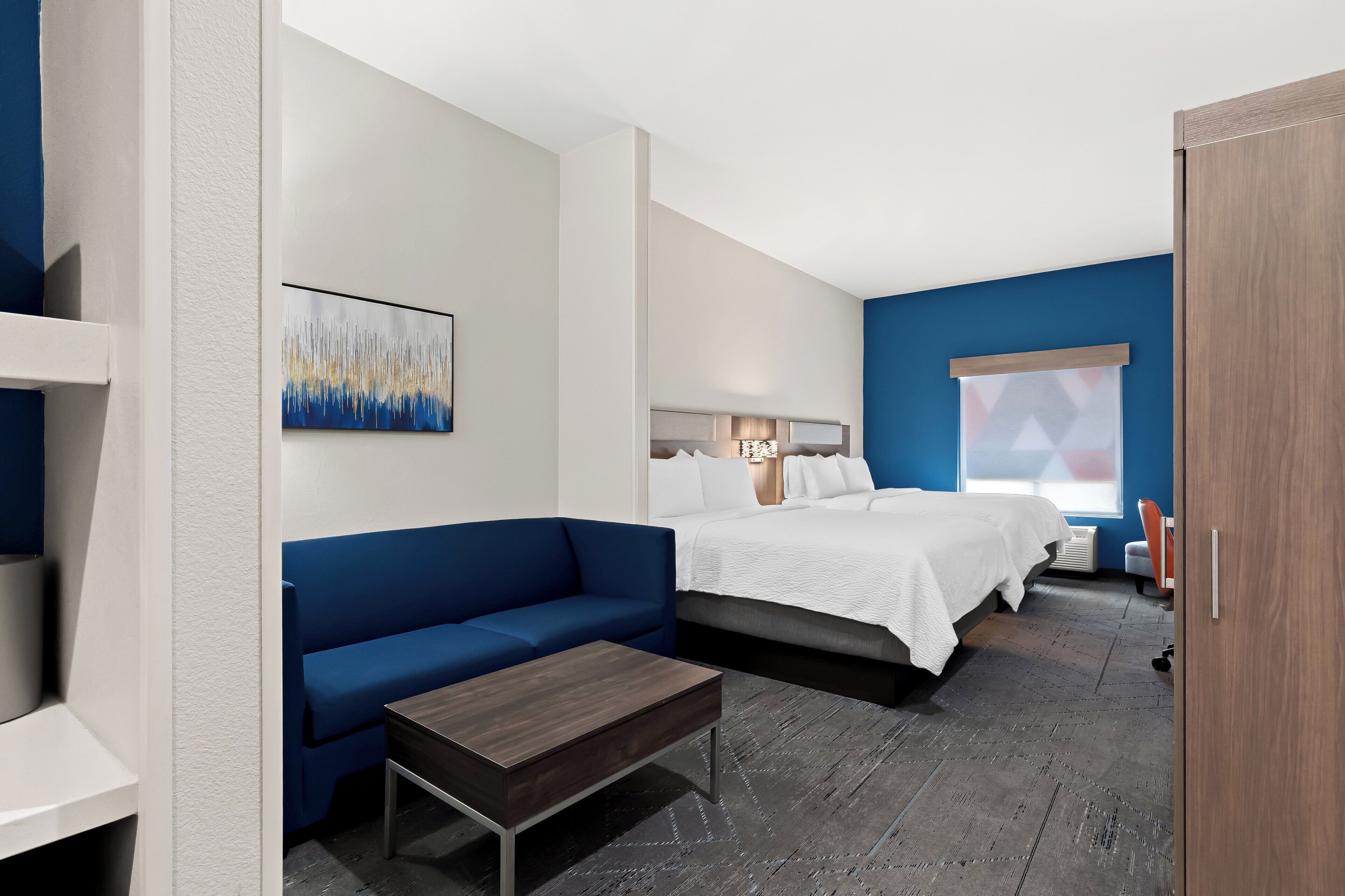 Quarto de hotel moderno com parede azul de destaque, cama branca, sofá azul e mesa de centro de madeira.