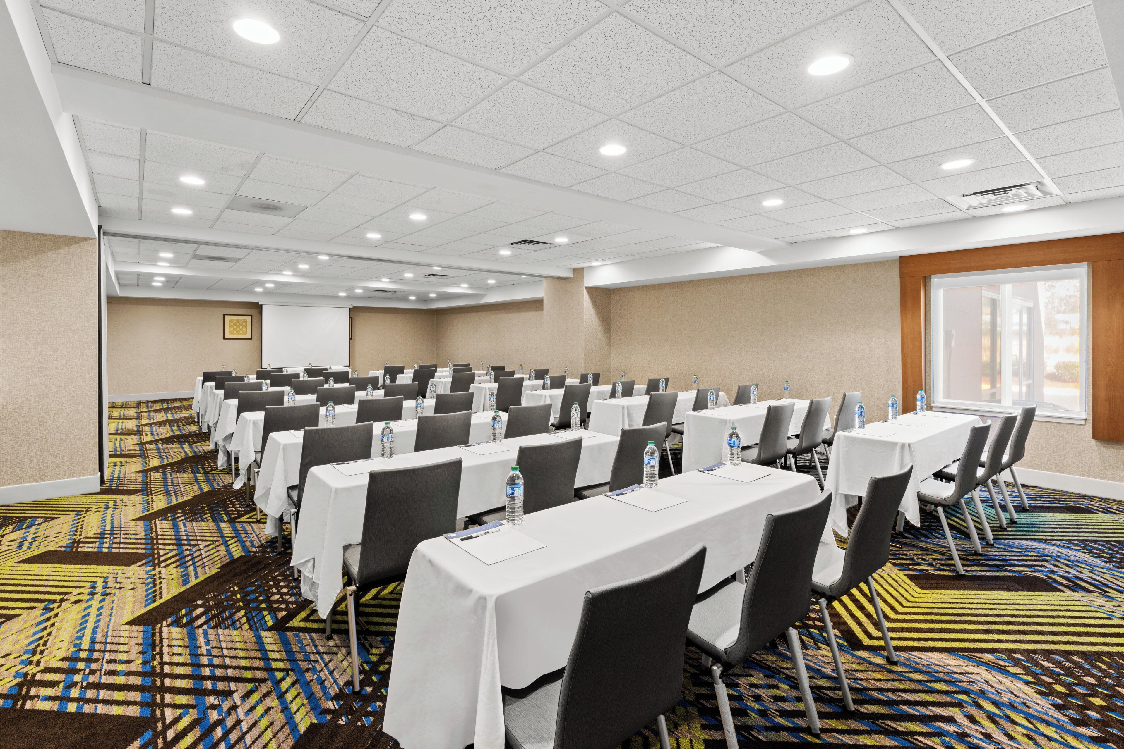 Sala de conferências com várias mesas e cadeiras, uma tela de projetor e um carpete estampado.