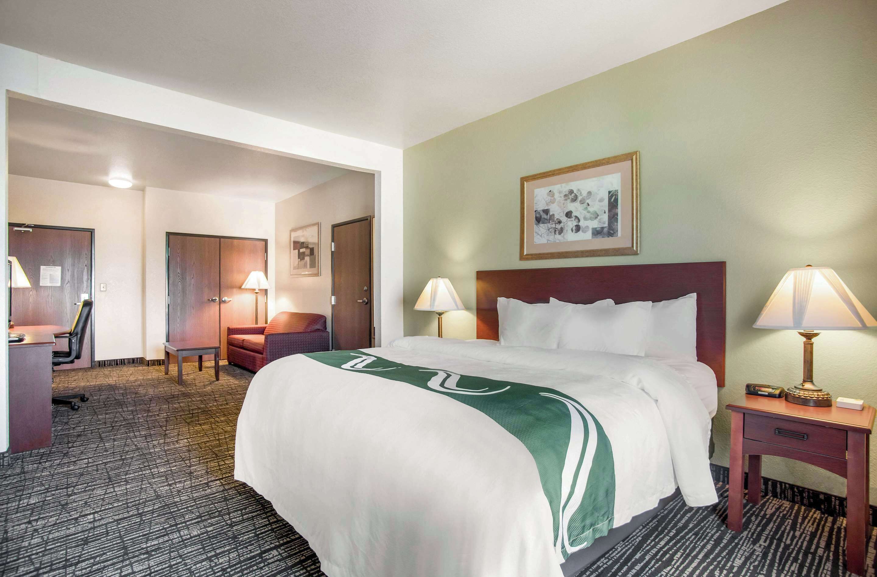 Quarto de hotel com uma cama grande, uma escrivaninha, uma cadeira, uma mesinha de cabeceira com uma luminária e um quadro na parede.