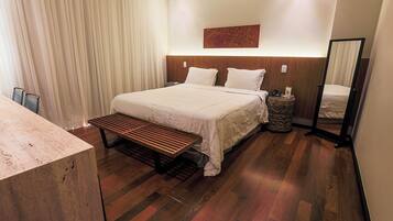 Quarto de hotel com uma cama grande, um banco de madeira, uma mesinha de cabeceira, um espelho e uma obra de arte pendurada na parede.