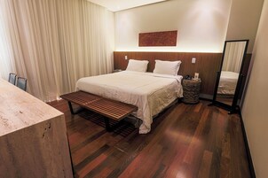 Quarto de hotel com uma cama grande, um banco de madeira, uma mesinha de cabeceira, um espelho e uma obra de arte pendurada na parede.