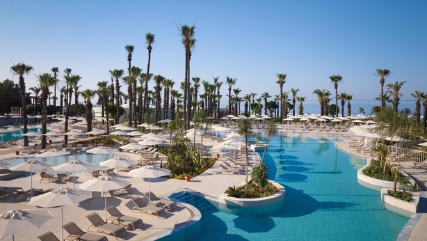 Área de piscina do resort com várias espreguiçadeiras e guarda-sóis, cercada por palmeiras e uma vista desobstruída do mar.