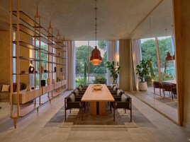 Uma sala de jantar moderna com uma longa mesa de madeira, várias cadeiras, uma estante e grandes janelas com vista para o verde.