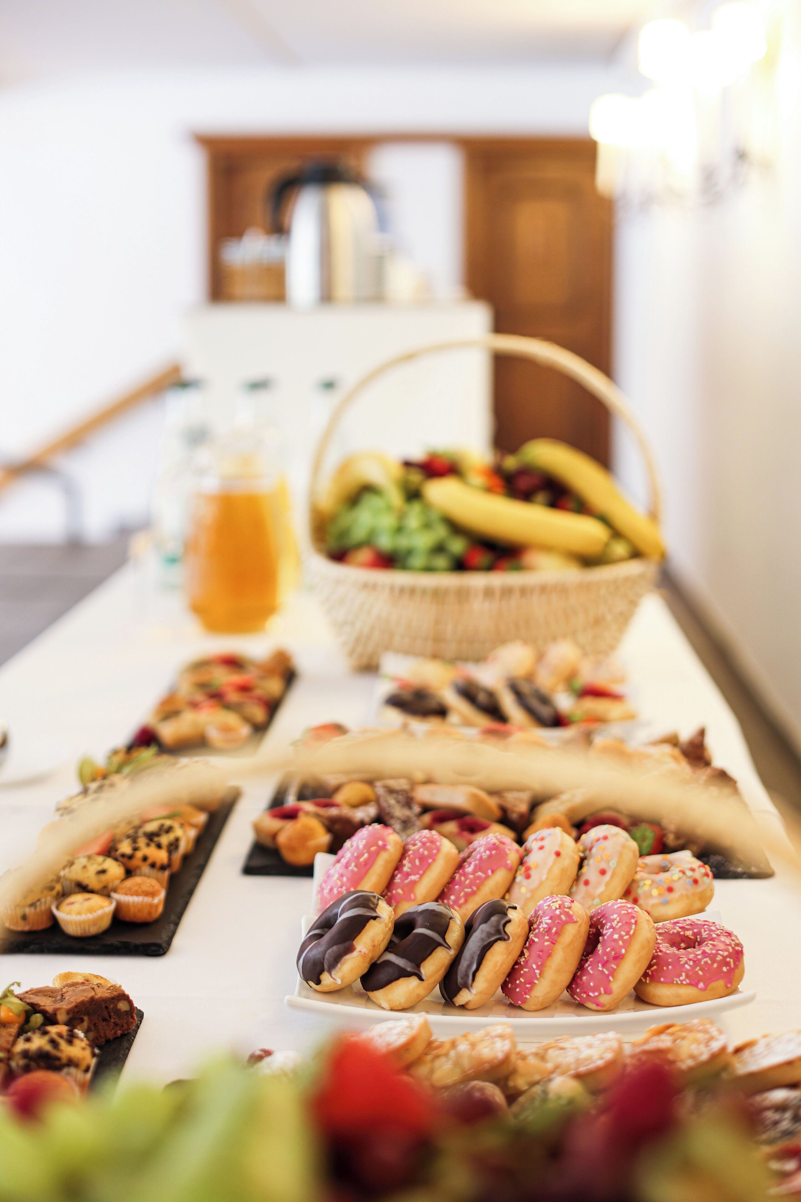 Uma mesa de buffet com diversos doces, uma cesta de frutas e um jarro de suco.