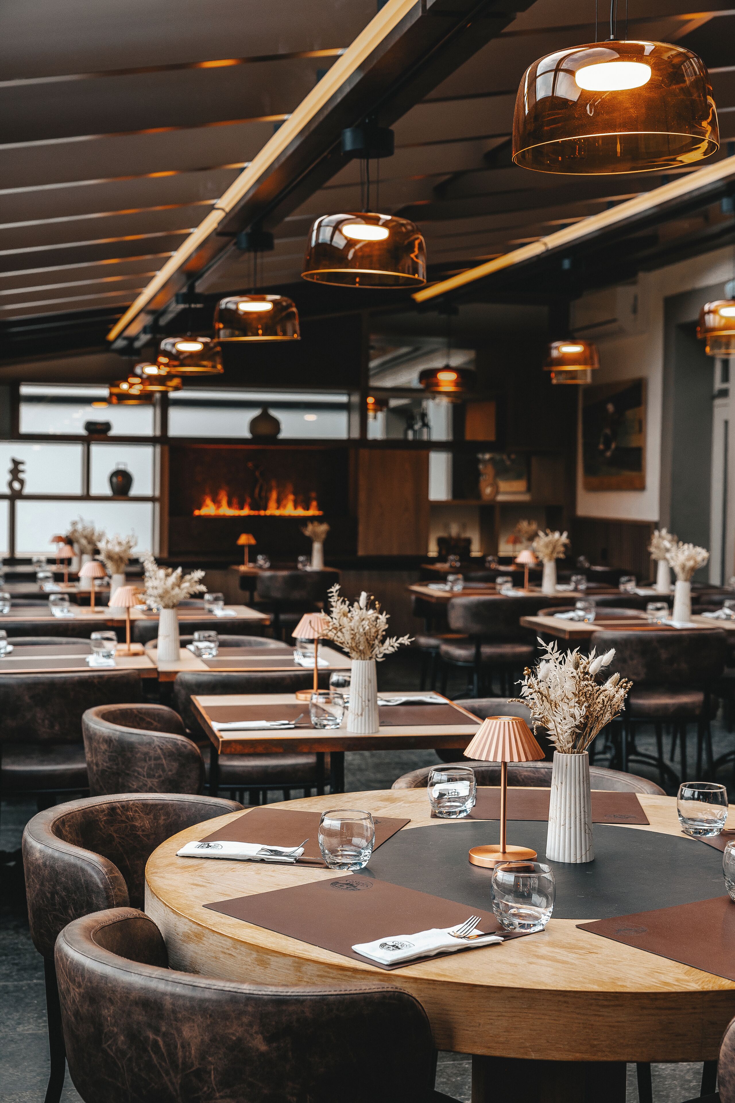 Um restaurante com mesas redondas, cadeiras de couro marrom e luminárias pendentes.