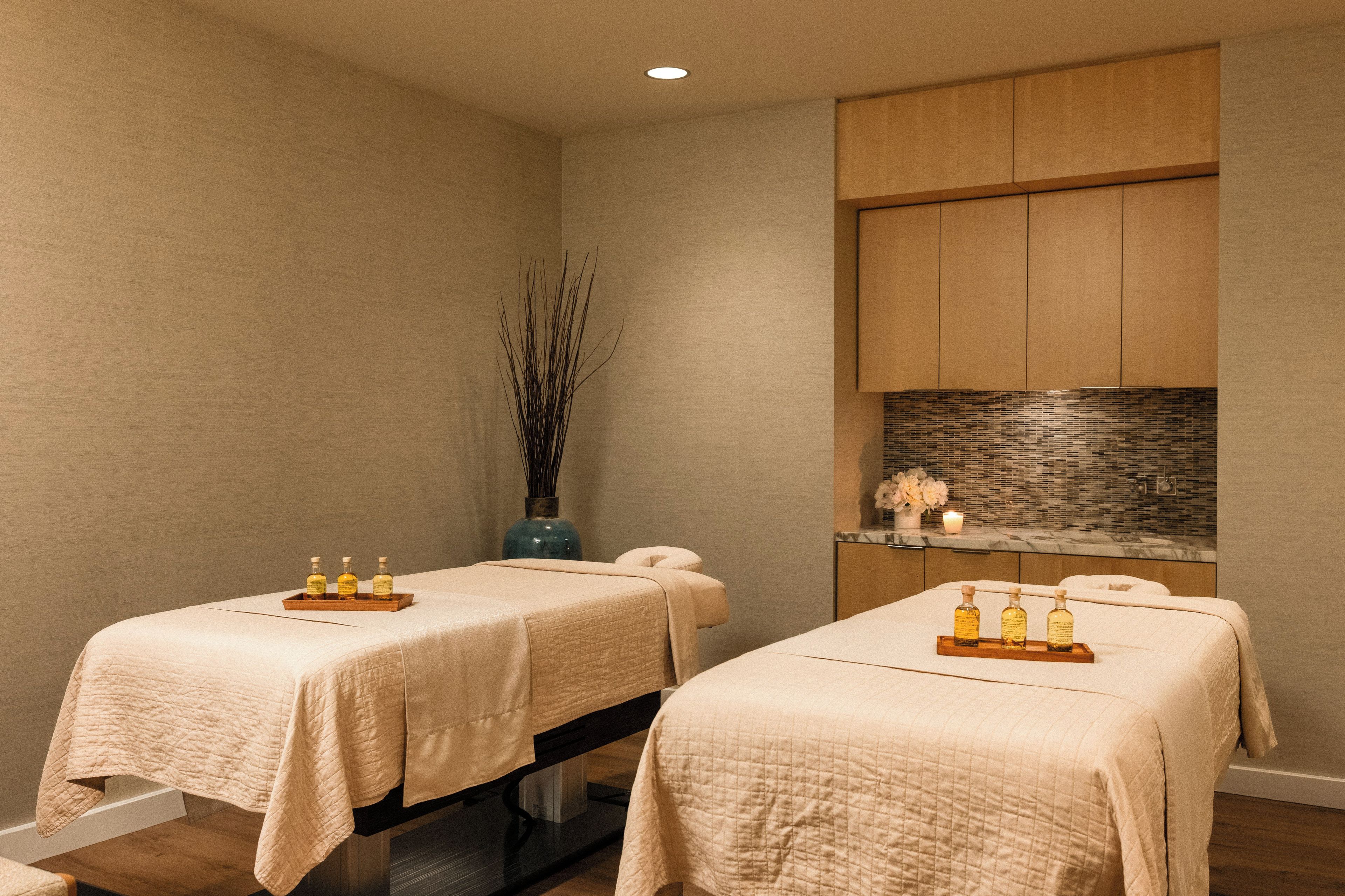 Sala de spa com duas mesas de massagem, cada uma com um tabuleiro de garrafas e um vaso com galhos.