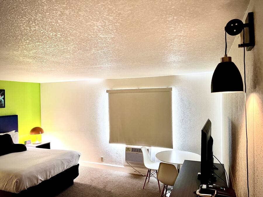 Quarto com cama, escrivaninha, cadeira, televisão e janela com persianas.