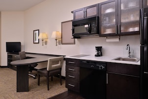 Quarto de hotel compacto com cozinha compacta, incluindo micro-ondas, cafeteira e pia. Há uma mesa com cadeira e televisão de tela plana.