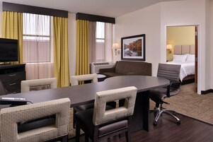 Quarto de hotel com uma grande mesa de conferência, quatro cadeiras, uma televisão, ar condicionado, uma cama e um quadro na parede.