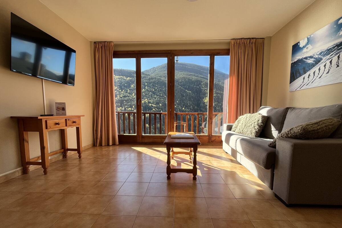 Sala de estar com sofá, mesa de madeira, TV na parede e varanda com vista para as montanhas.