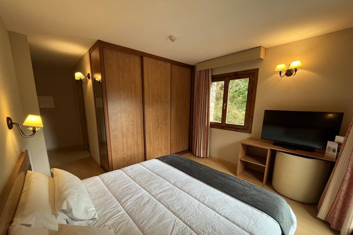 Quarto com cama, televisão, guarda-roupa e janela com cortinas.