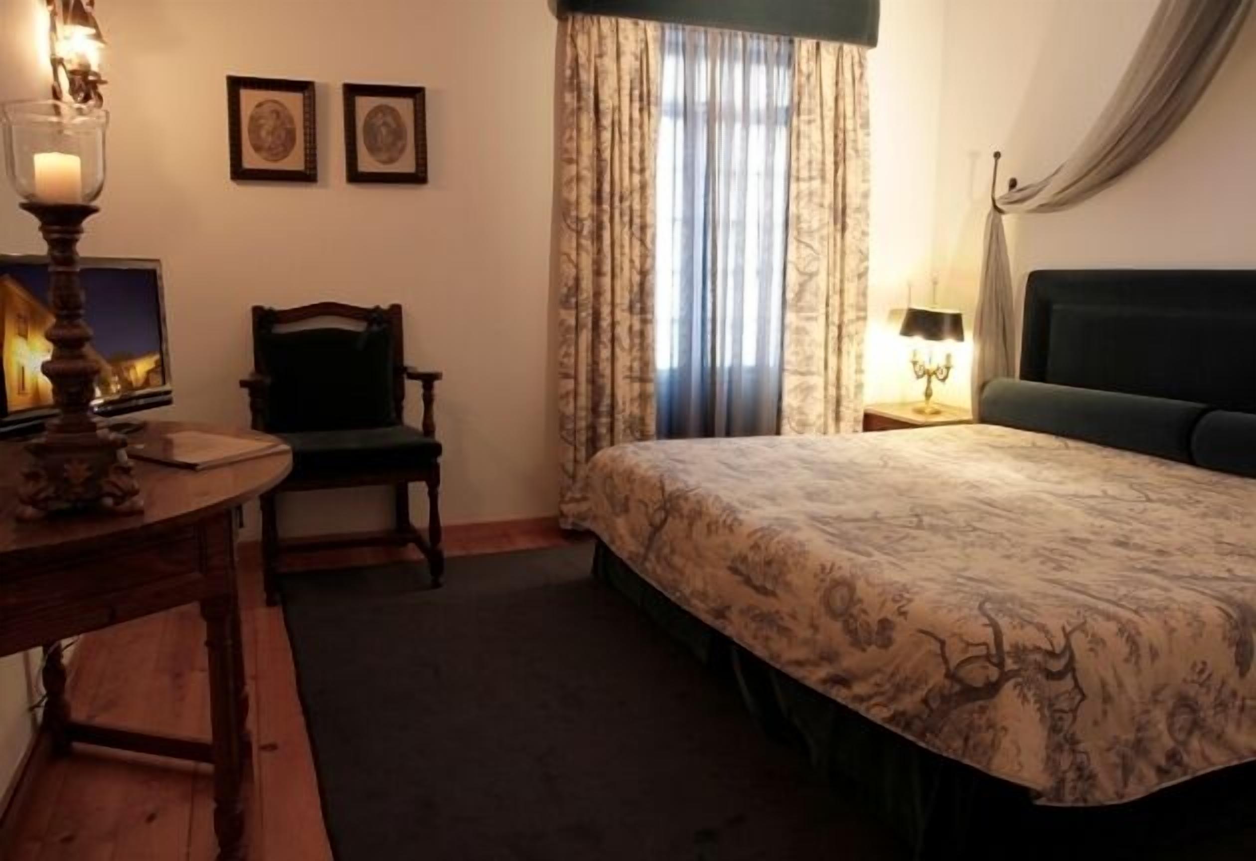 Quarto de hotel com cama, escrivaninha, cadeira, luminária e cortinas.
