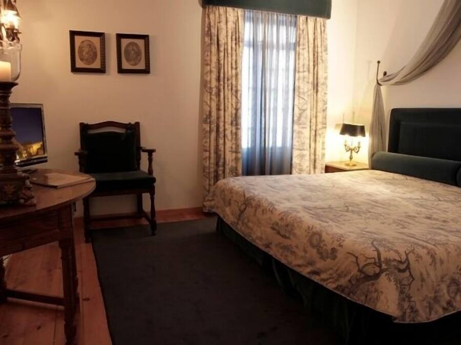 Quarto de hotel com cama, escrivaninha, cadeira, luminária e cortinas.