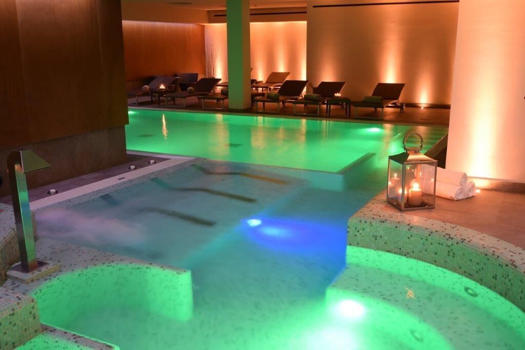 Piscina interior com iluminação verde, espreguiçadeiras e piso revestido de azulejos.