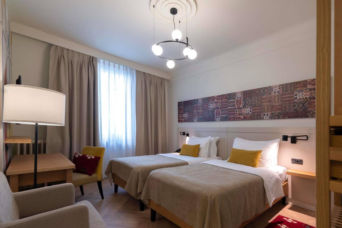 Quarto de hotel com duas camas, uma escrivaninha, uma cadeira, um abajur e uma grande pintura abstrata na parede.