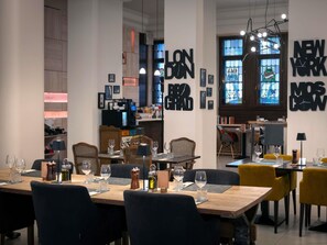 Uma área de refeições moderna com mesas para dois, um canto para café e uma grande obra de arte na parede retratando nomes de cidades.