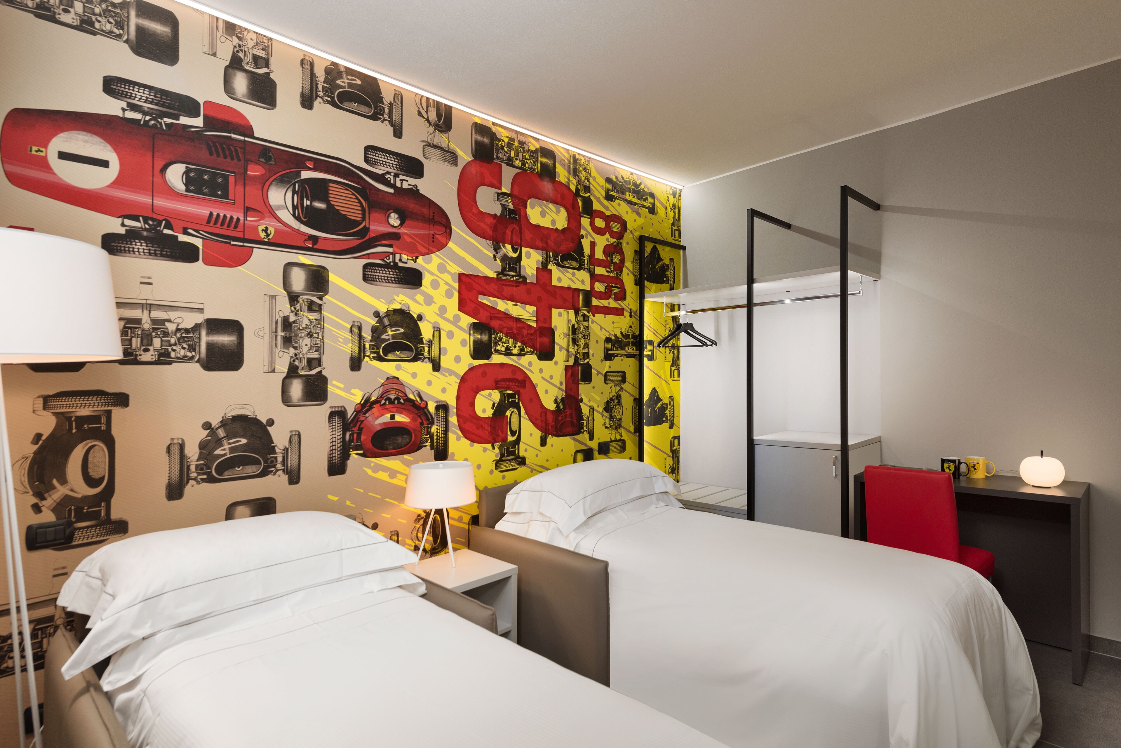 Quarto de hotel com duas camas, um grande mural na parede com carros de corrida, um espelho, uma cadeira vermelha e uma mesa de cabeceira com abajur.