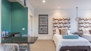Quarto de hotel moderno com cozinha revestida em azulejos verde-água, cama com cabeceira estampada e obra de arte na parede listando diversos locais.