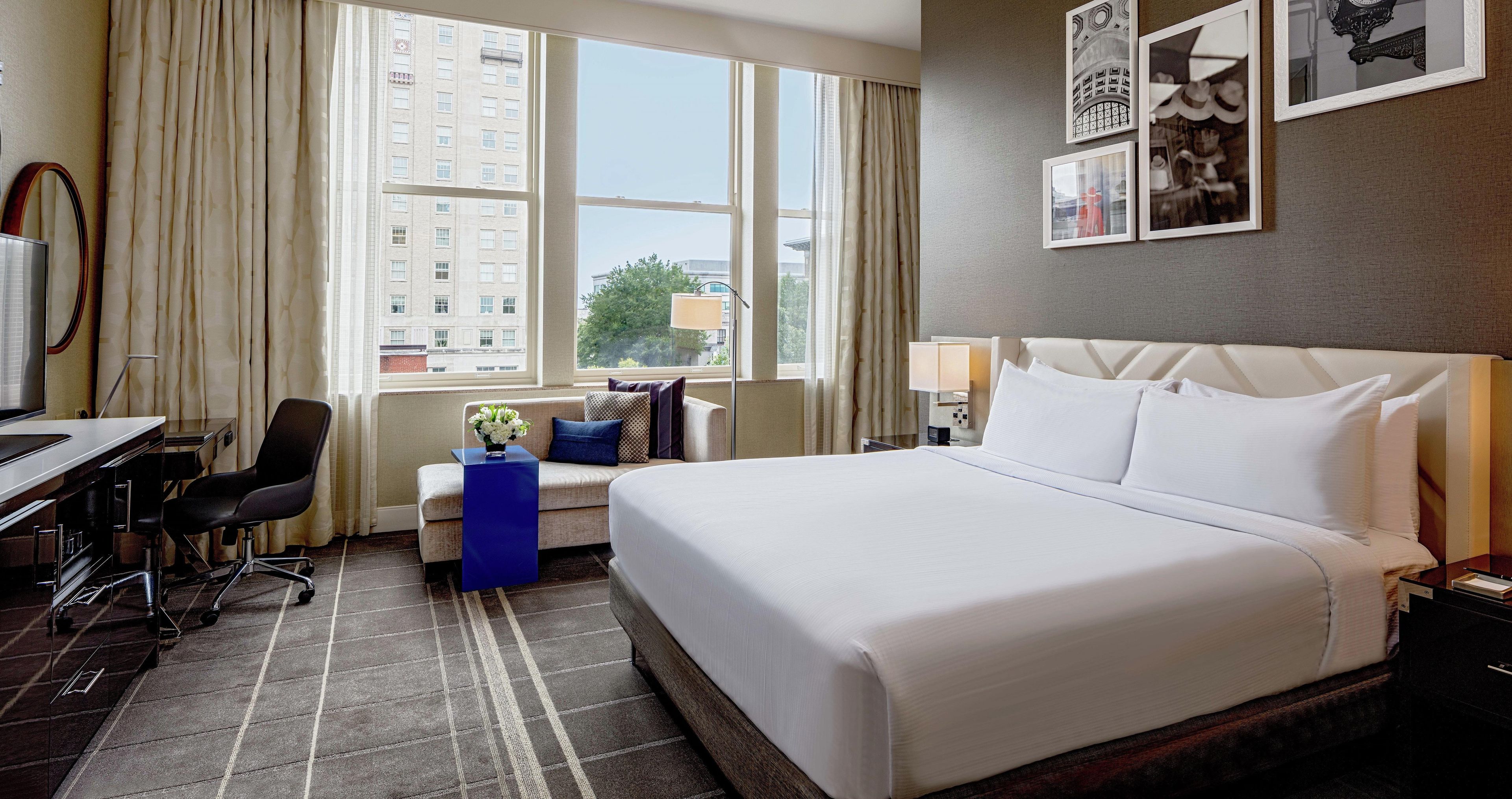 Quarto de hotel com uma cama grande, uma mesa e uma cadeira. Há uma janela com vista para edifícios e árvores.