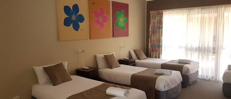 Quarto de hotel com três camas, uma janela grande com cortinas translúcidas e três quadros de flores na parede.