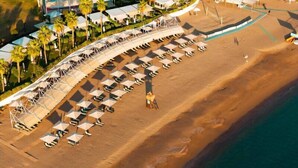 Vista aérea de uma praia com fileiras de espreguiçadeiras e guarda-sóis, uma piscina e palmeiras.