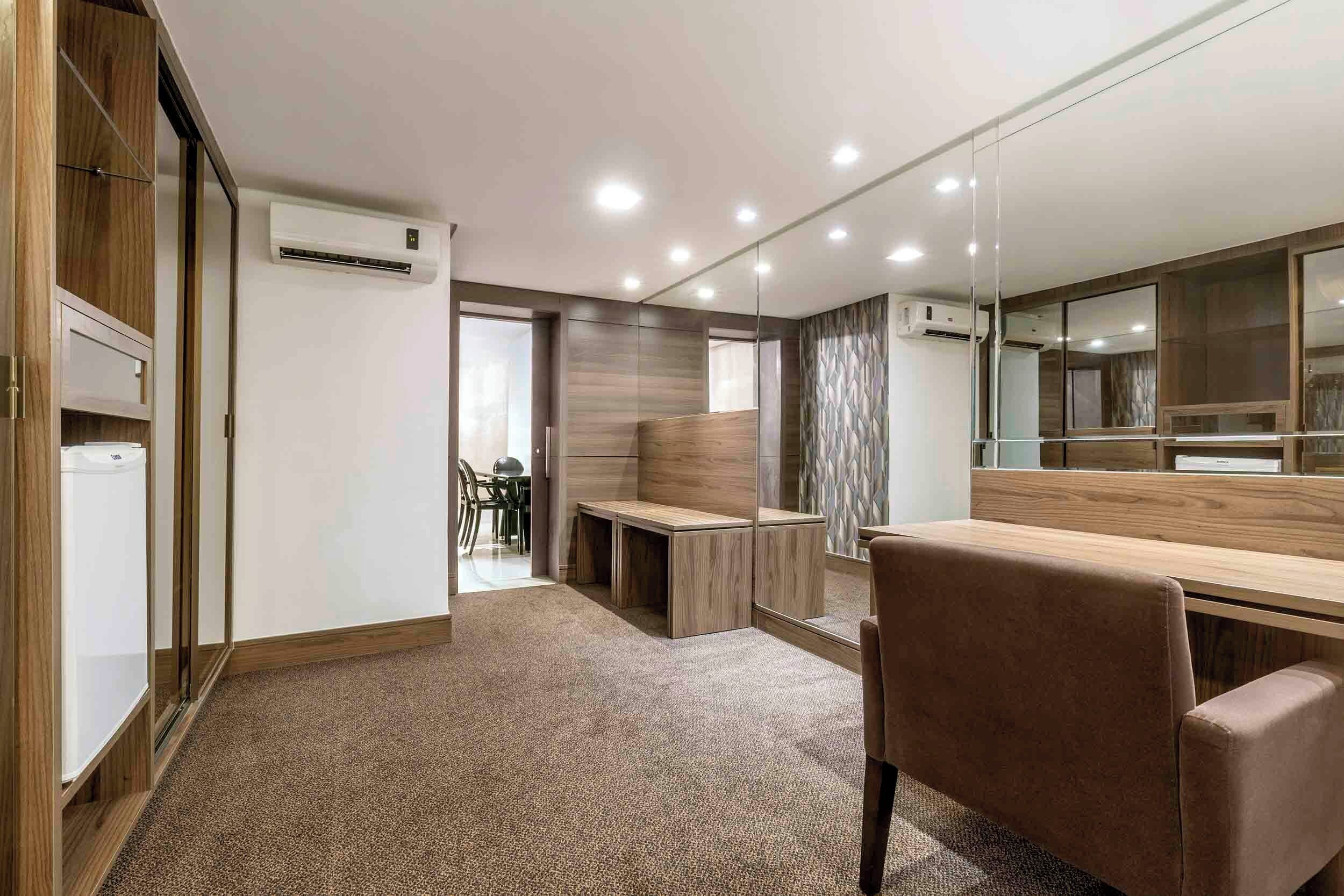 Quarto moderno com escrivaninha de madeira, uma cadeira marrom, um banco e um espelho grande.