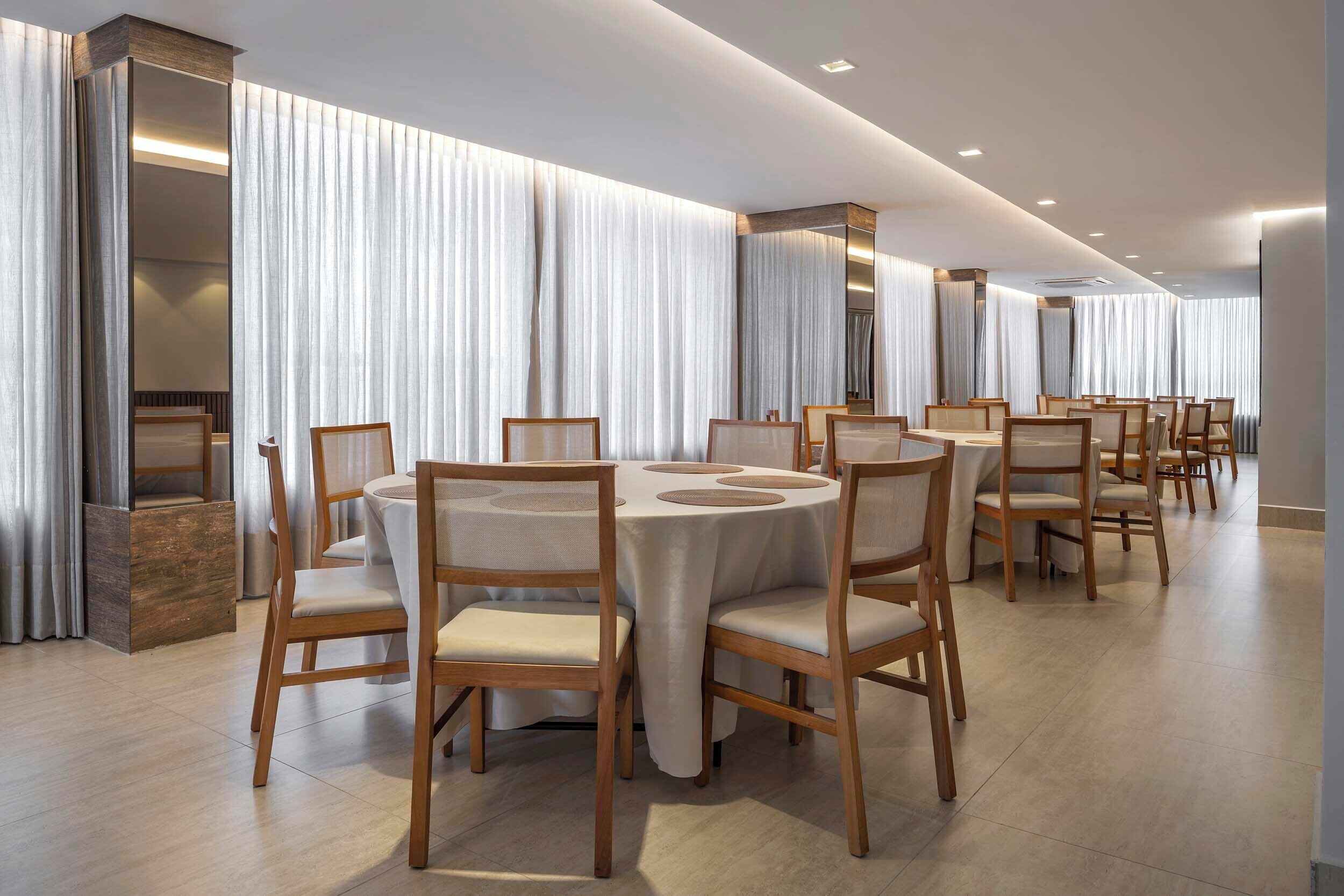 Uma sala de jantar moderna com mesas redondas, cadeiras de madeira e uma janela grande com cortinas.