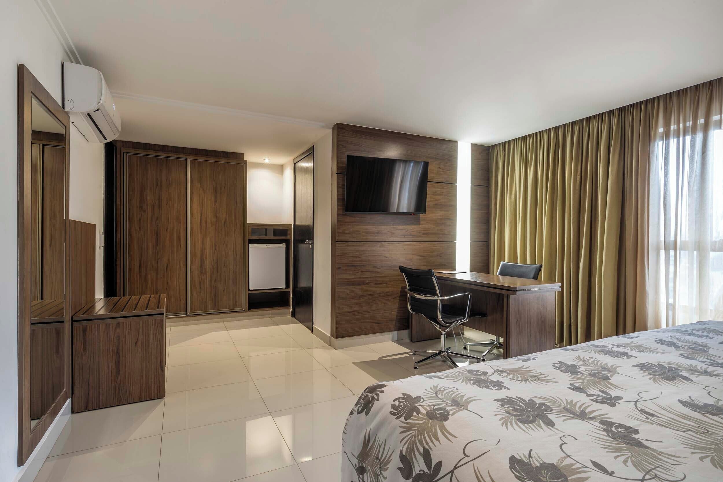 Quarto de hotel com cama, escrivaninha, cadeira, televisão, guarda-roupa e ar condicionado.