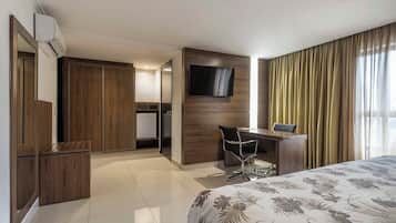 Quarto de hotel com cama, escrivaninha, cadeira, televisão, guarda-roupa e ar condicionado.