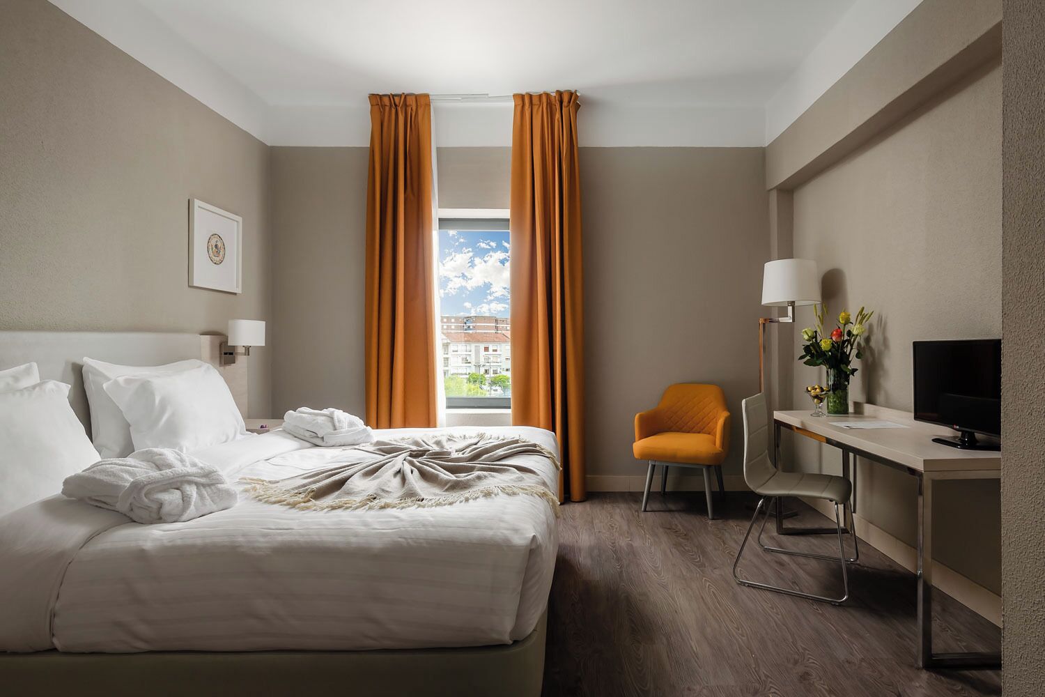 Quarto de hotel com cama, escrivaninha com cadeira, televisão e janela com cortinas.