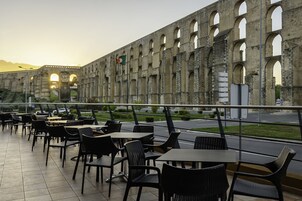Um café ao ar livre com mesas e cadeiras, com vista para um edifício histórico com arcos e uma bandeira.