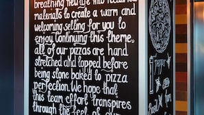 Um quadro negro com texto manuscrito descrevendo o processo de preparo da pizza e o ambiente acolhedor do local.
