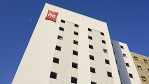 Um edifício moderno com um destaque para o letreiro 'ibis' na fachada, apresentando uma grade de janelas e um exterior branco e limpo.