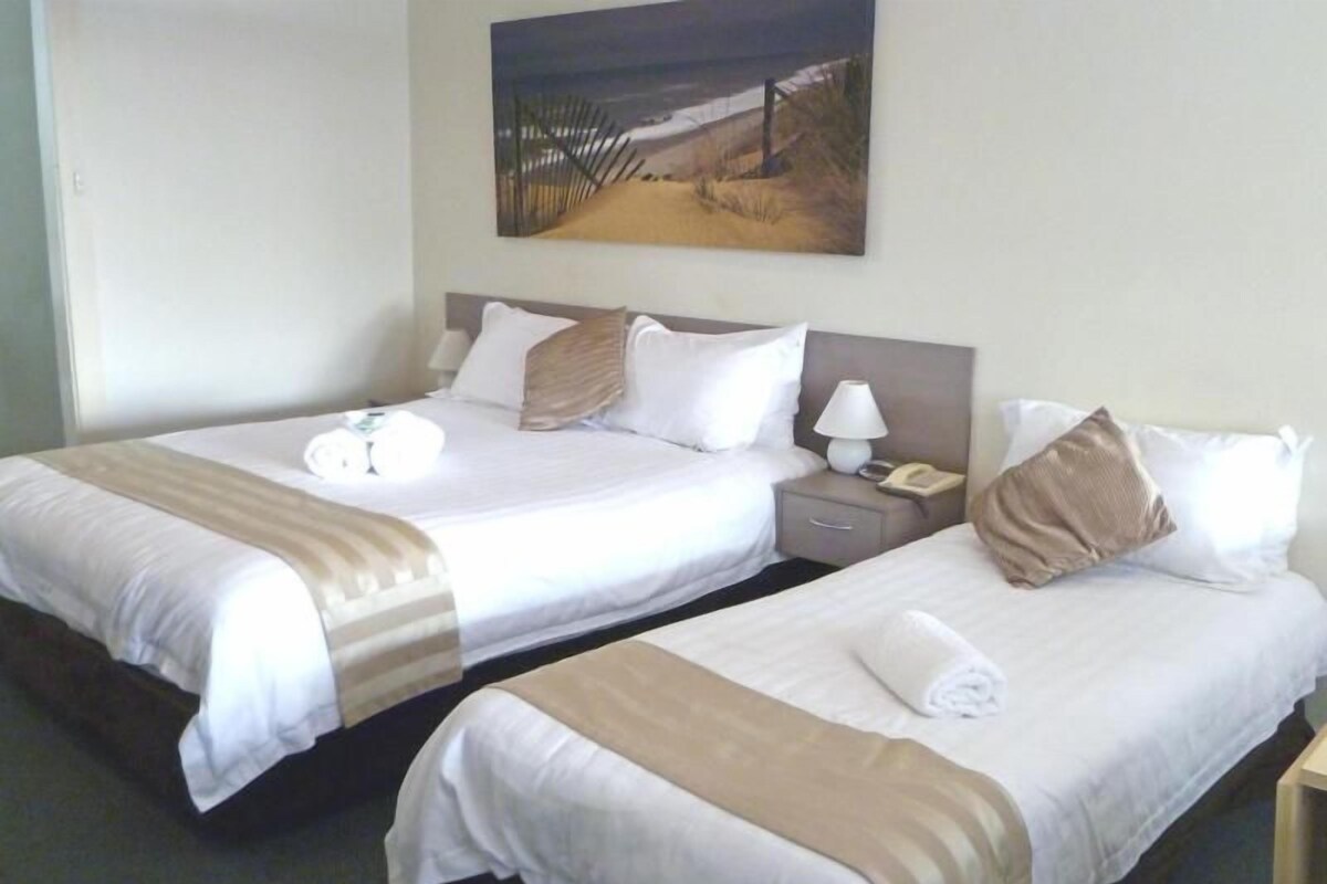 Quarto de hotel com duas camas, cada uma com roupa de cama branca e detalhes bege, uma mesa de cabeceira com abajur, um quadro na parede e vista para a praia.