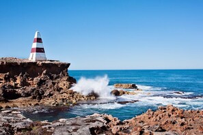 Um litoral rochoso com um farol em um penhasco, ondas quebrando nas rochas e um céu azul límpido.