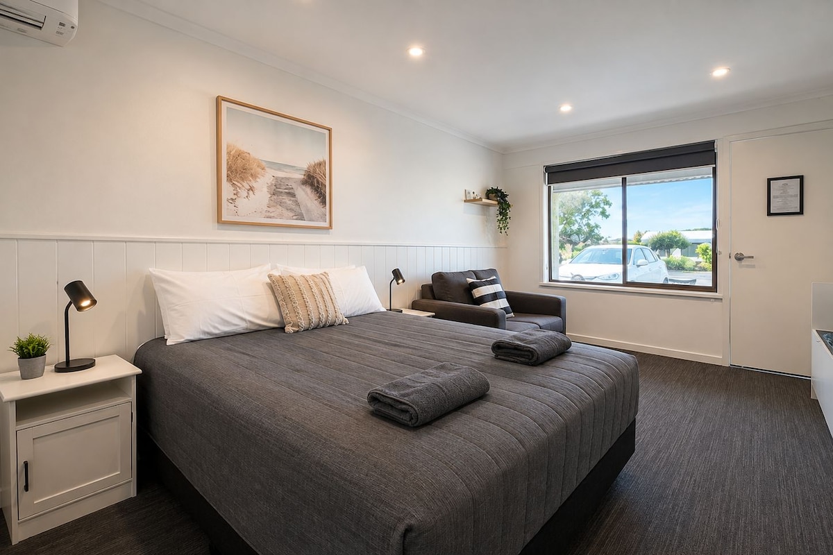 Quarto com cama grande, mesas de cabeceira, escrivaninha, cadeira, janela com vista e um quadro na parede.