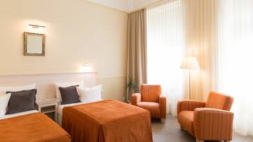 Quarto de hotel com duas camas, uma cadeira, um abajur e uma janela grande com cortinas.