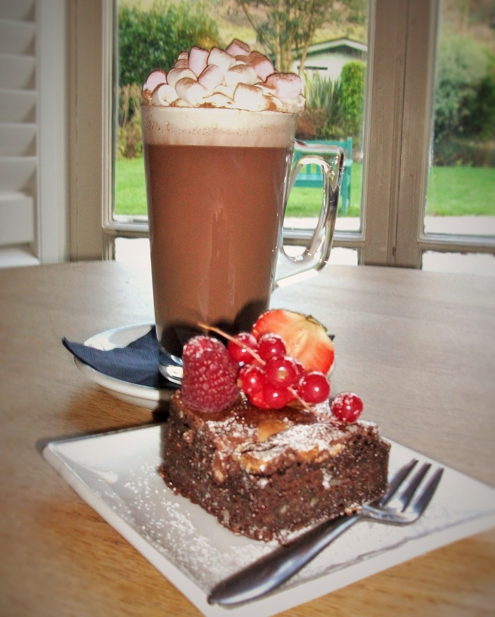 Sobremesa com fatia de bolo coberta com frutas vermelhas e açúcar em pó, um copo de chocolate quente e um garfo sobre o prato.