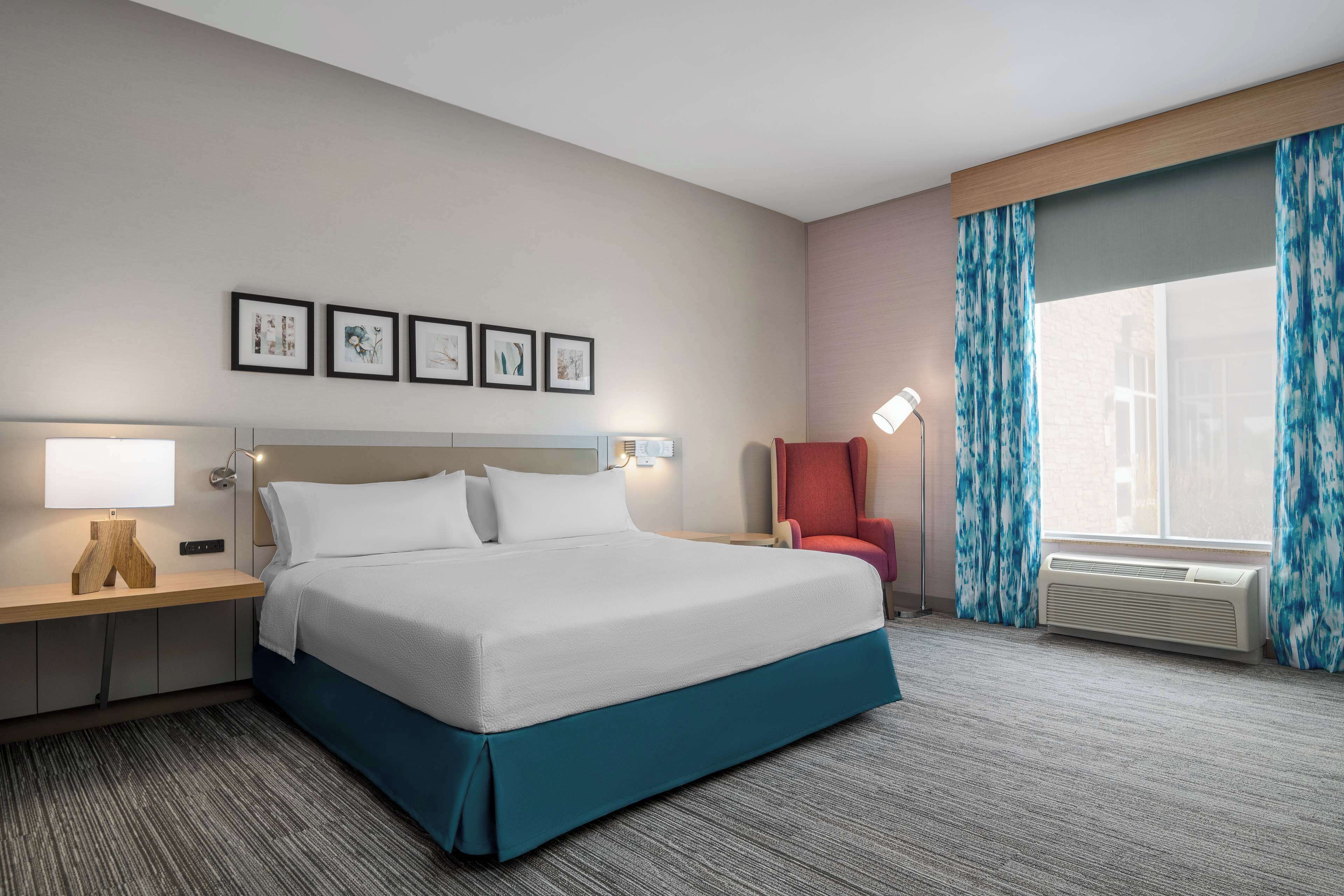 Quarto de hotel com uma cama grande, uma escrivaninha com luminária, uma cadeira e uma janela com cortinas.