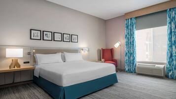 Quarto de hotel com uma cama grande, uma escrivaninha com luminária, uma cadeira e uma janela com cortinas.