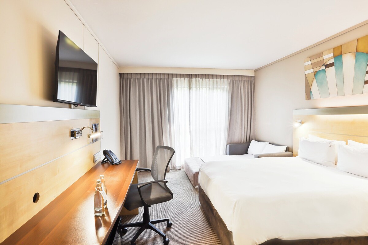 Quarto de hotel com cama, escrivaninha com telefone, cadeira, televisão fixada na parede e janela com cortinas.