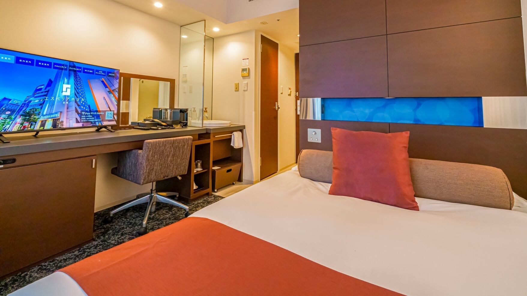 Quarto de hotel com cama, escrivaninha, cadeira e uma televisão grande exibindo uma vista urbana.