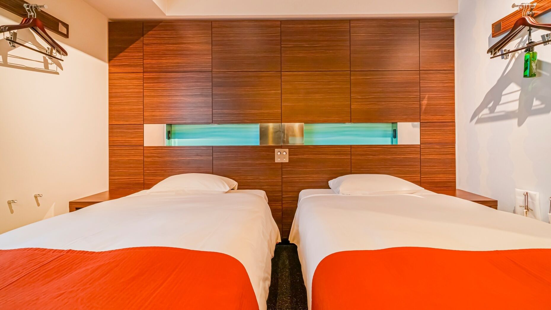 Quarto de hotel com duas camas, cabeceira de madeira, prateleira embutida e cabideiro.