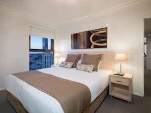 Quarto de hotel com uma cama grande, duas mesinhas de cabeceira, uma janela com vista para a cidade e um quadro na parede.