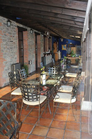 Um pátio externo com uma mesa comprida e cadeiras, cercado por paredes de pedra e vigas de madeira.
