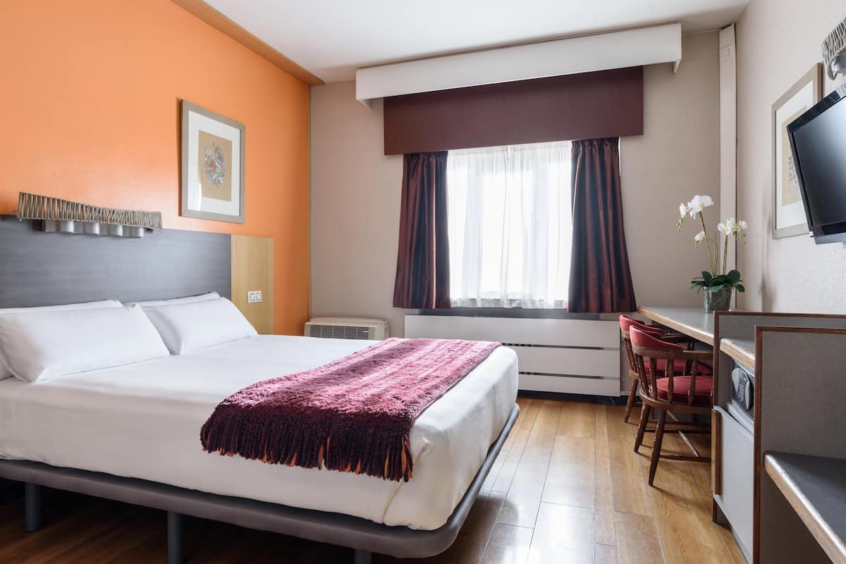 Quarto de hotel com cama, escrivaninha, cadeira, televisão e janela com cortinas.