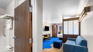 Quarto de hotel com porta de madeira, sofá azul, escrivaninha com cadeira, televisão de tela plana e uma janela grande.