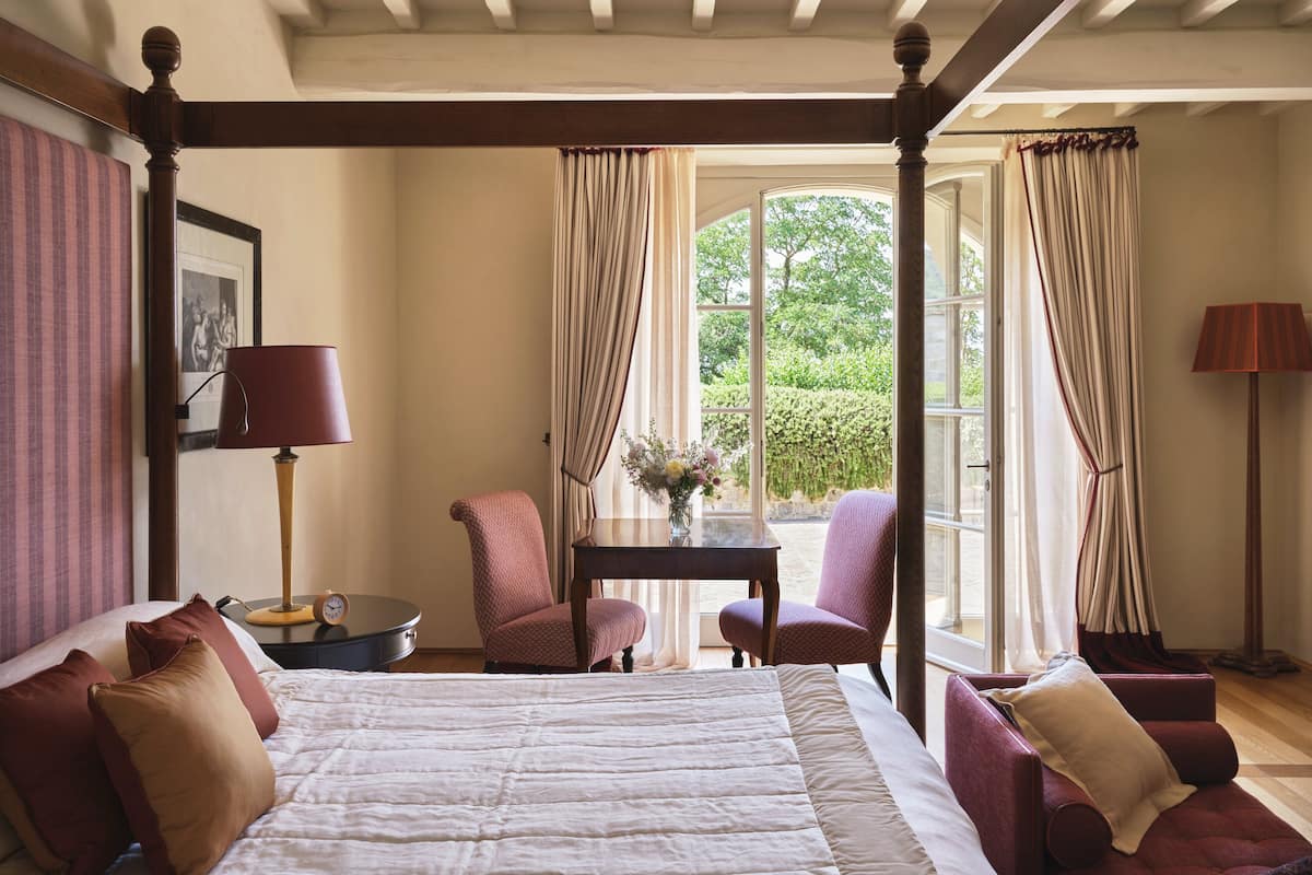Quarto com uma cama grande, duas cadeiras, uma mesa e uma luminária. Há uma janela com cortinas e vista para o verde lá fora.