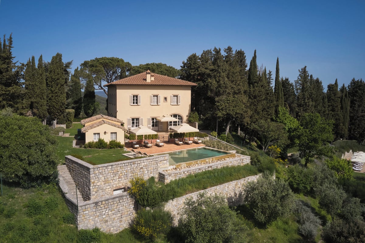 Uma grande casa de dois andares com piscina, cercada por vegetação exuberante e árvores altas.