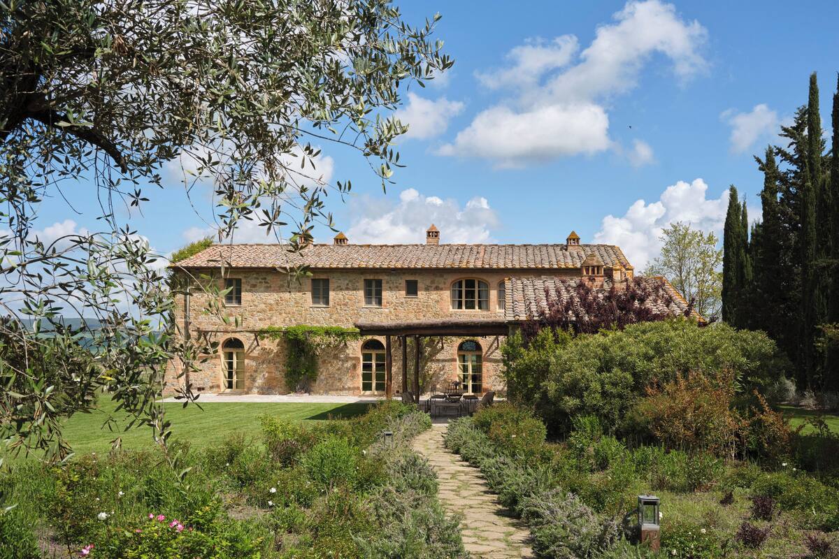Uma casa de pedra com telhado de cerâmica, um caminho de pedra que leva à entrada e um jardim com diversas plantas e flores.